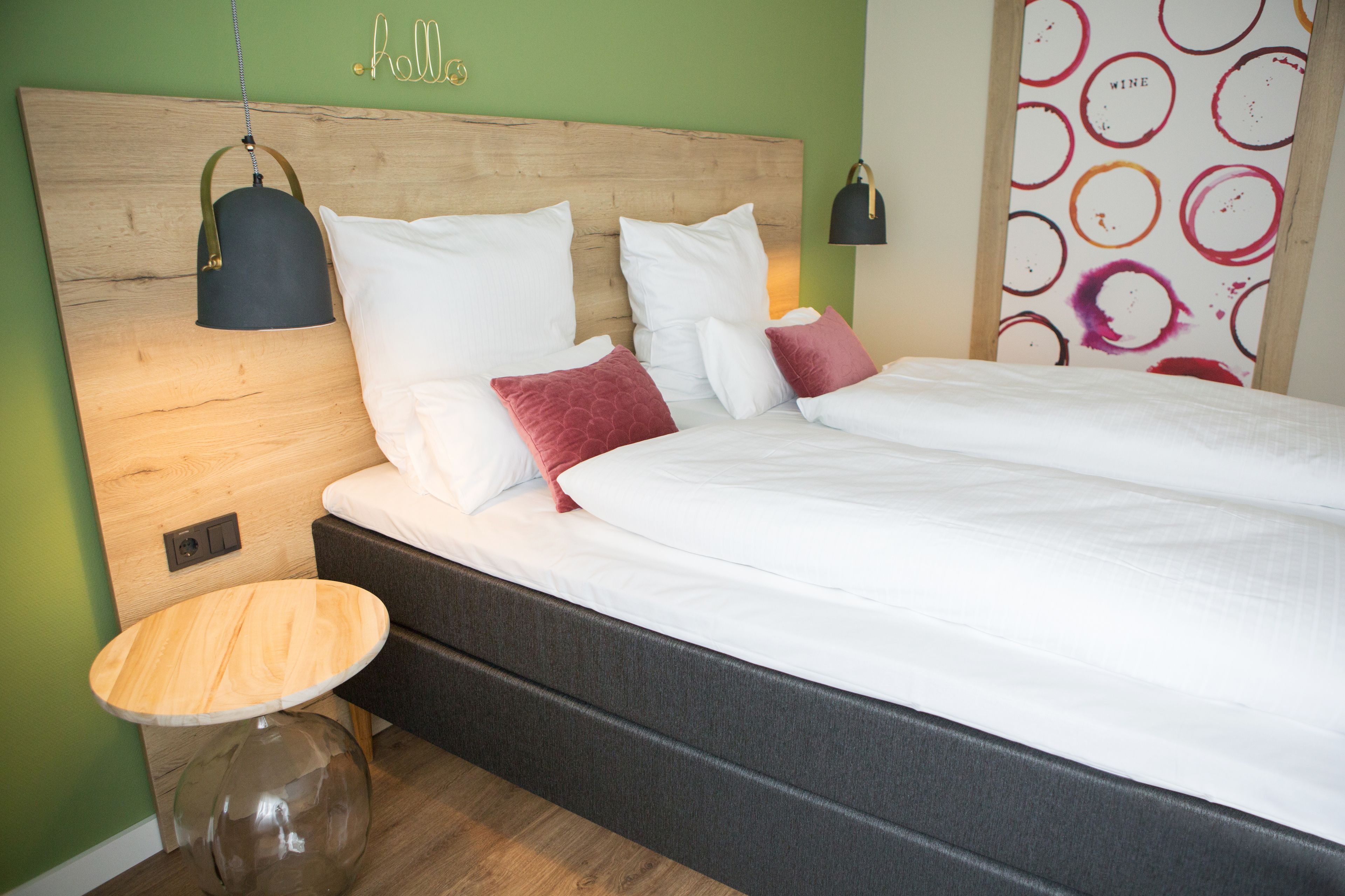 Cama bem arrumada com almofadas brancas e vermelhas, cabeceira de madeira e uma pequena mesa lateral.