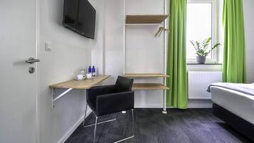 Quarto de hotel com cama, escrivaninha com cadeira, televisão na parede, janela com cortinas verdes e uma planta em vaso no parapeito.