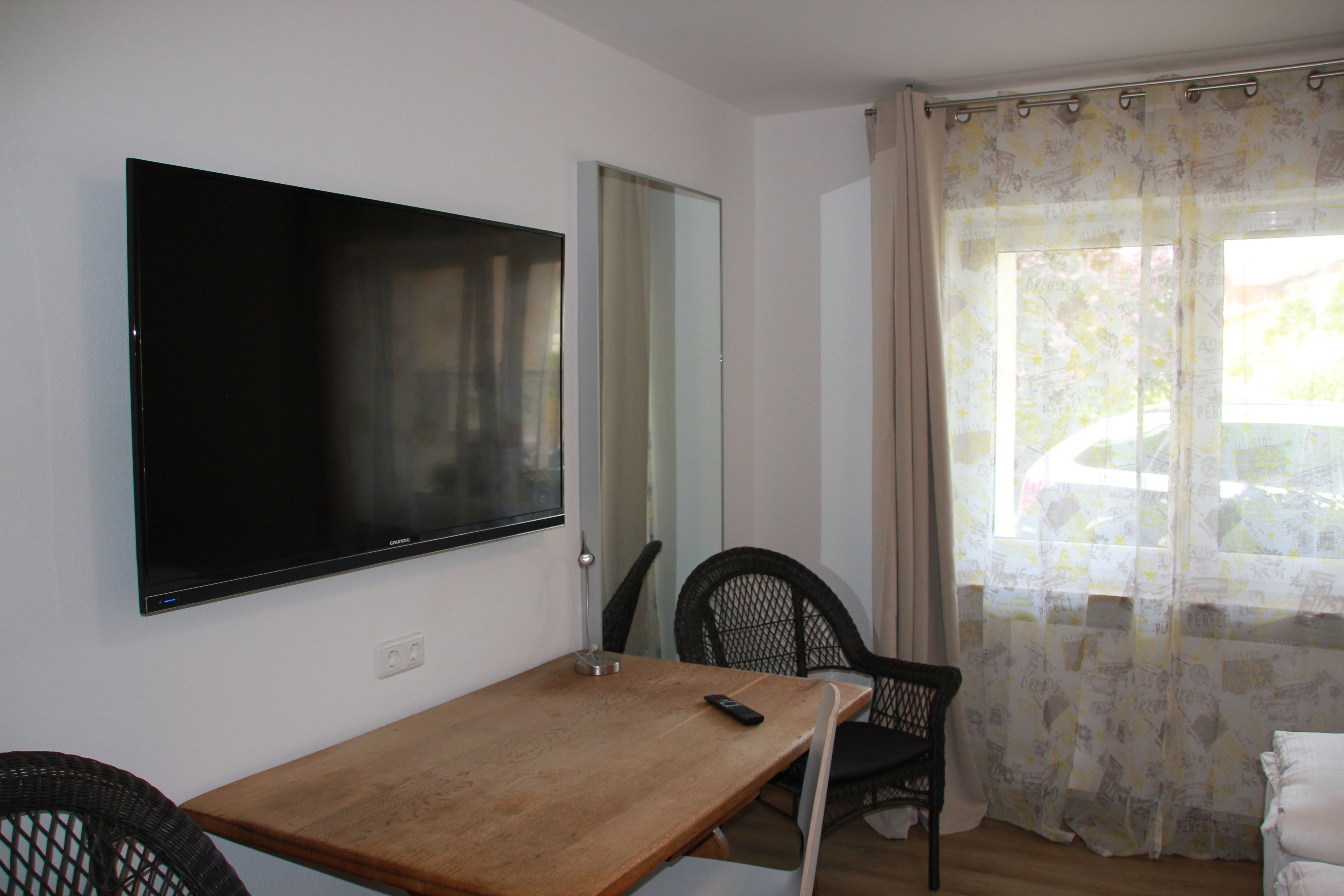 Quarto com televisão de tela plana na parede, mesa de madeira com controle remoto, duas cadeiras de vime e janela com cortinas estampadas.