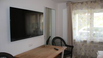 Quarto com televisão de tela plana na parede, mesa de madeira com controle remoto, duas cadeiras de vime e janela com cortinas estampadas.