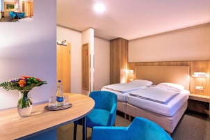 Quarto de hotel com duas camas, uma escrivaninha com um vaso de flores, uma garrafa e duas cadeiras azuis.