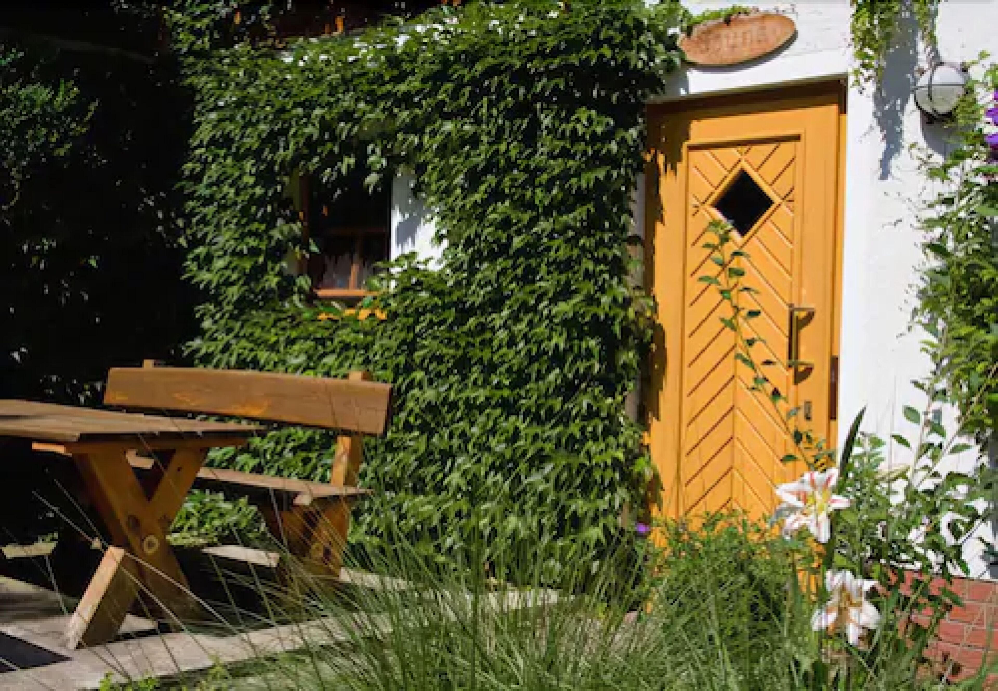 Um banco de madeira e uma porta com padrão geométrico em ziguezague, rodeados por vegetação.
