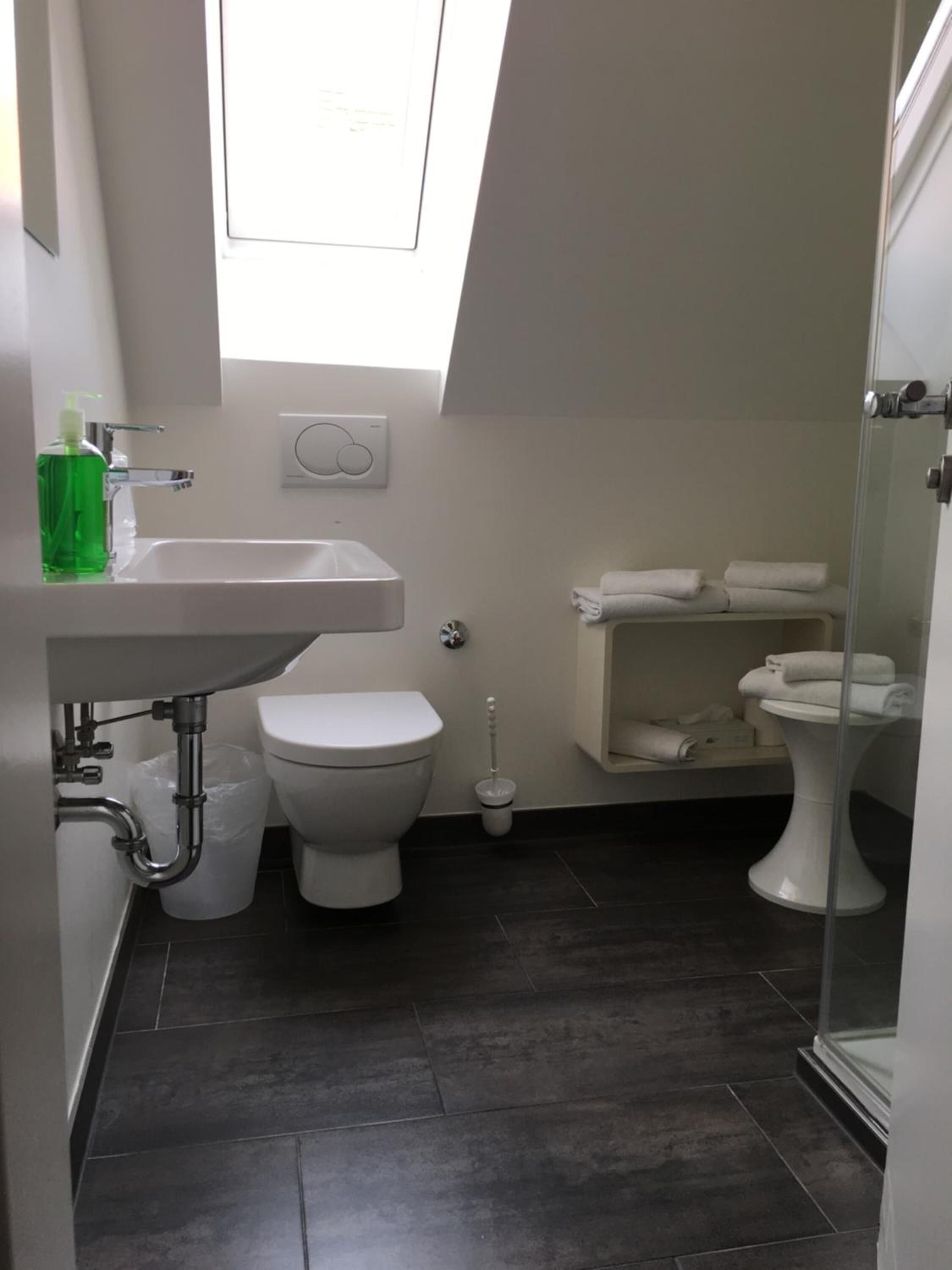 Casa de banho compacta com lavatório branco, sanita e bidé. Há uma janela sobre o lavatório e toalhas estão visíveis numa prateleira.