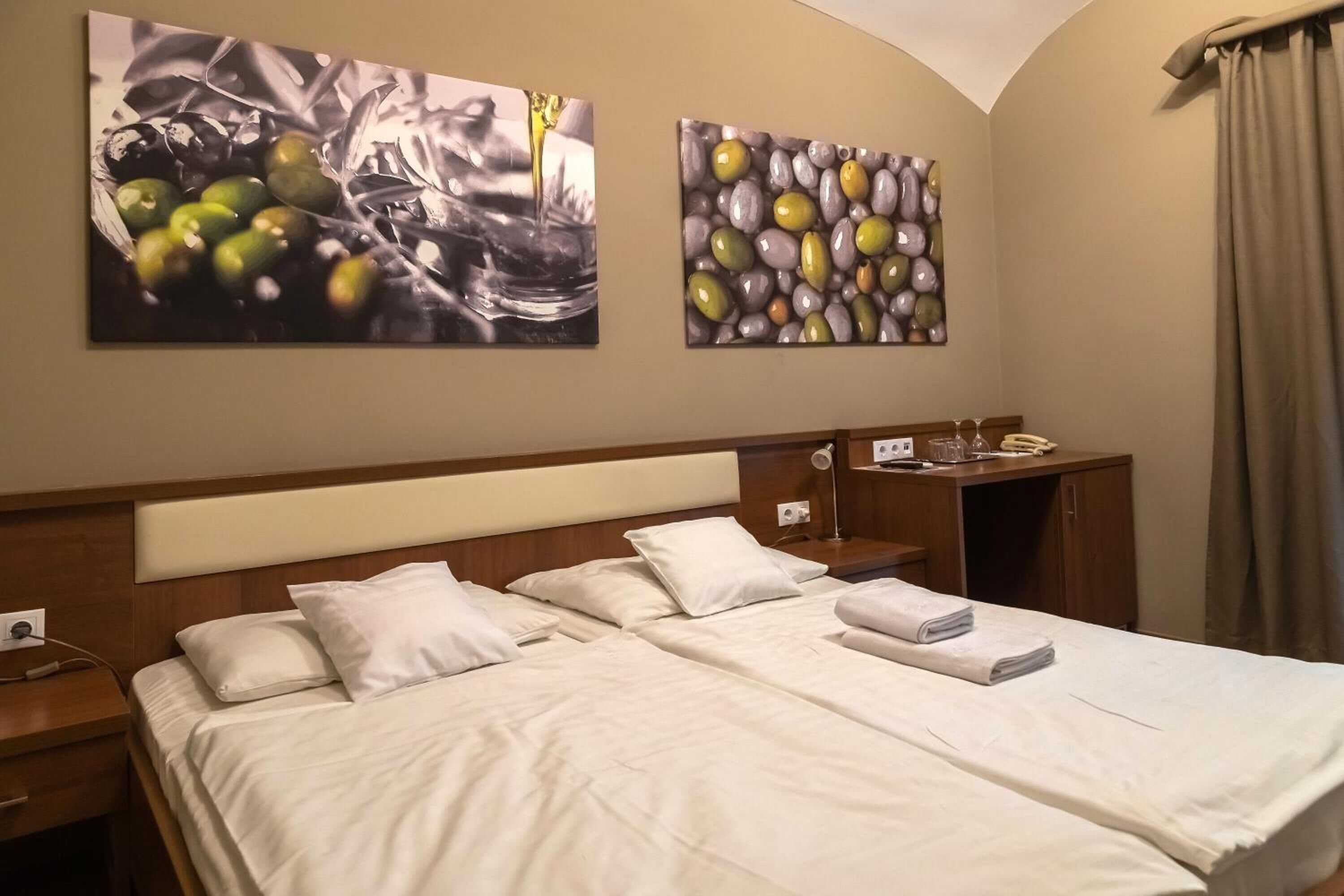 Quarto de hotel com uma cama grande, duas almofadas brancas, uma cabeceira de madeira, uma mesa de cabeceira com abajur e duas pinturas penduradas na parede.