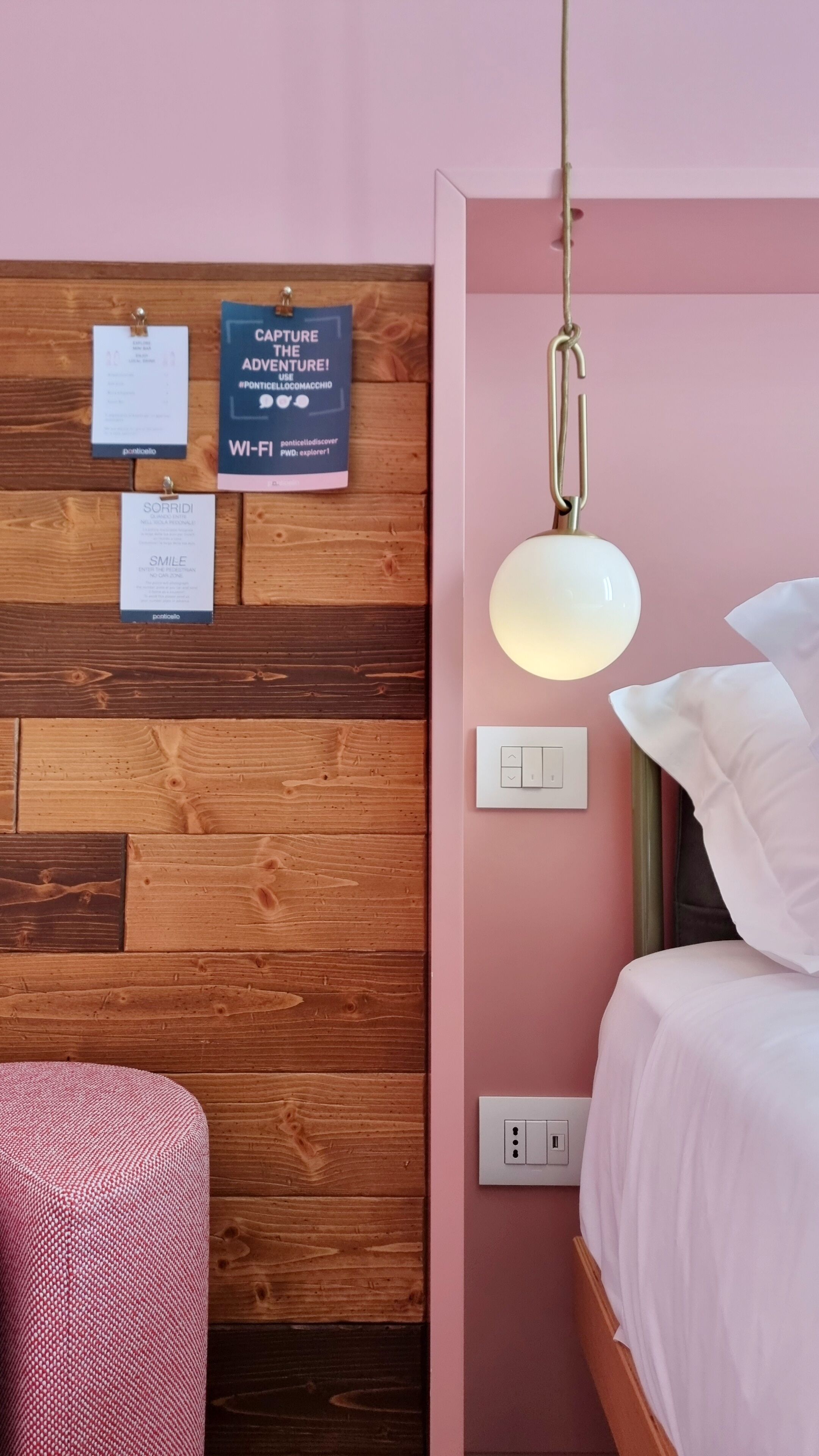 Quarto com painel de parede de madeira, cama com roupa de cama branca, luminária pendente e interruptor de luz fixado na parede.