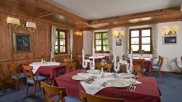 Uma sala de jantar com paredes revestidas de madeira, mesas com toalhas brancas e cadeiras azuis, e arandelas decorativas.