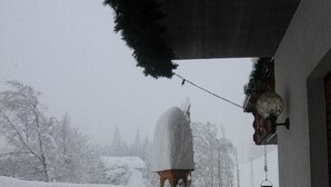 Uma área externa coberta de neve com um banco e uma cadeira de madeira, uma estátua com neve e um edifício com varanda.