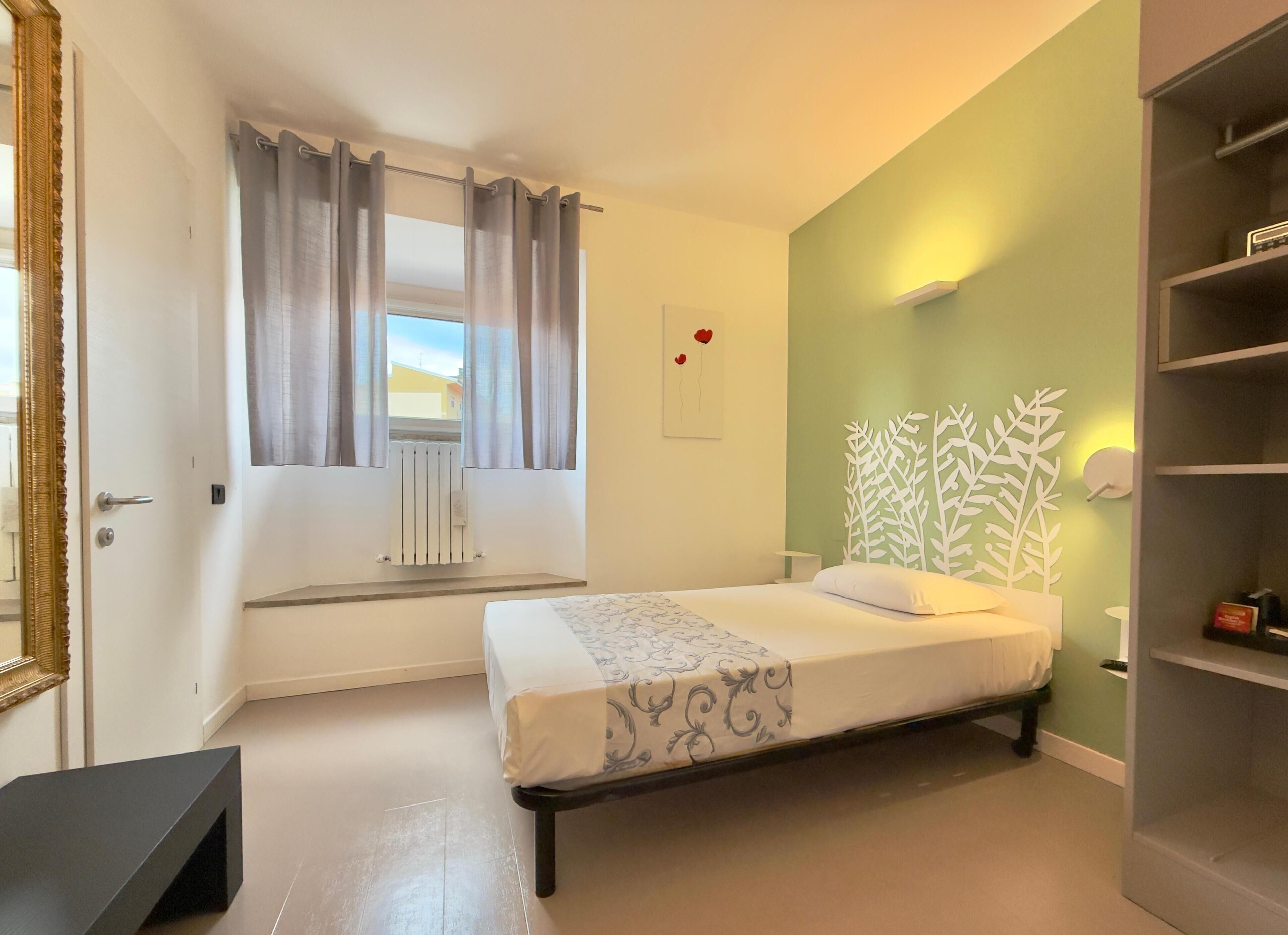 Quarto com cama, radiador, janela com cortinas, parede com um desenho de planta decorativa e uma estante com objetos.
