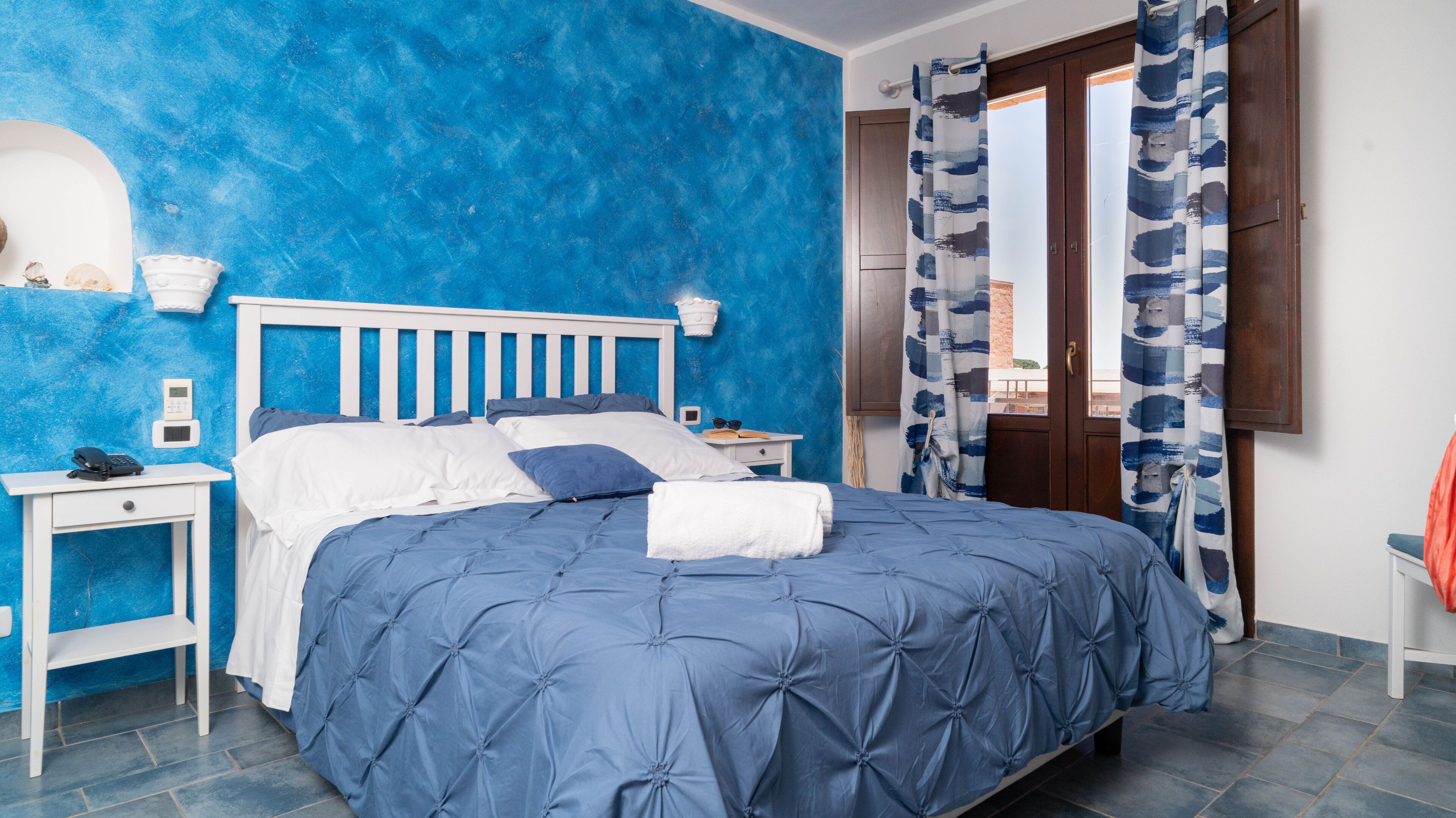 Quarto com parede azul texturizada, cama branca com roupa de cama azul, mesa de cabeceira com telefone e luminária, e janela com cortinas azuis.