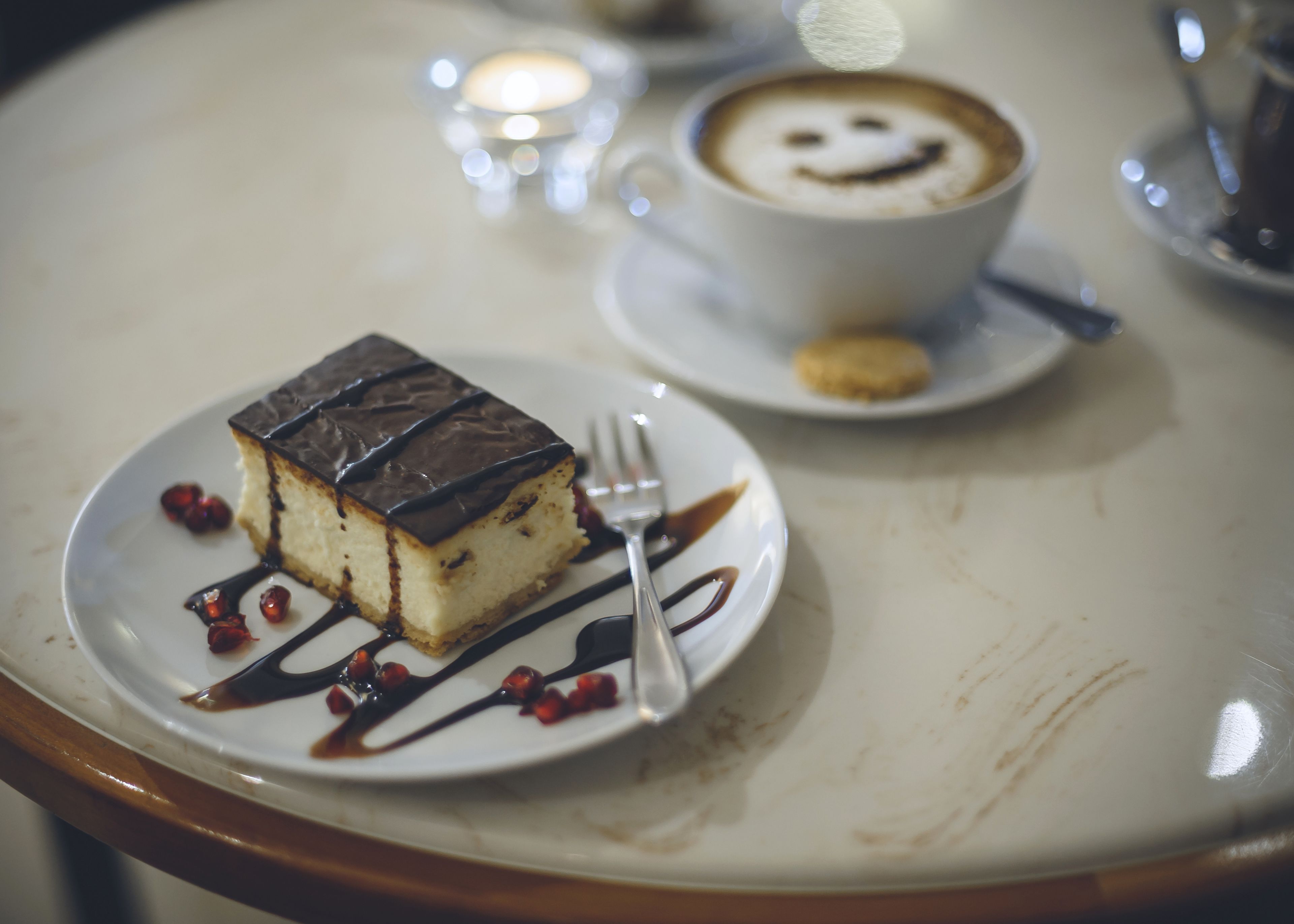 Um prato de sobremesa com uma fatia de bolo e calda de chocolate, uma xícara de café com um desenho sorridente no creme e um garfo sobre a mesa.