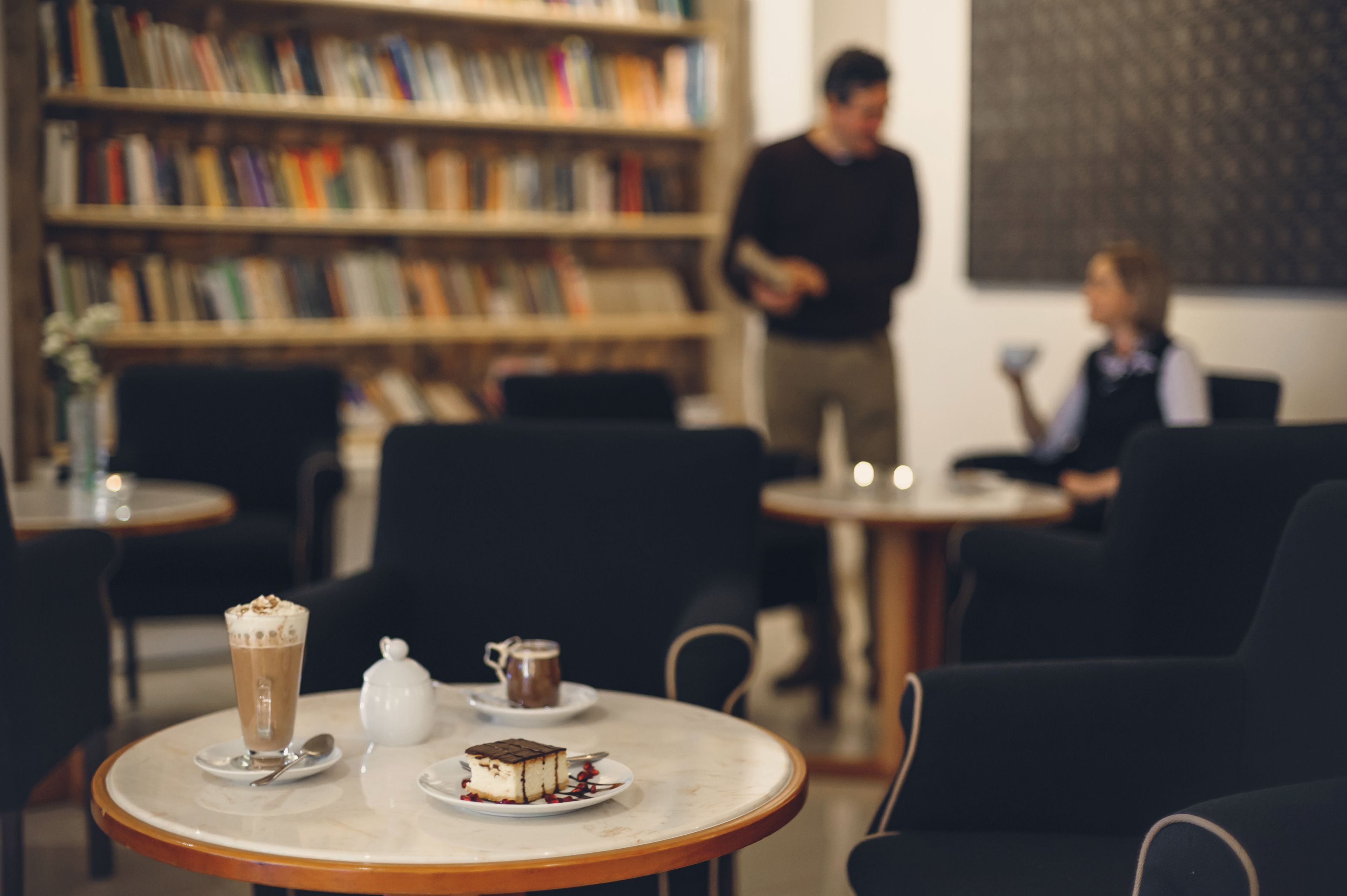 Um café acolhedor com mesas redondas, uma estante de livros e um muro com lousa.