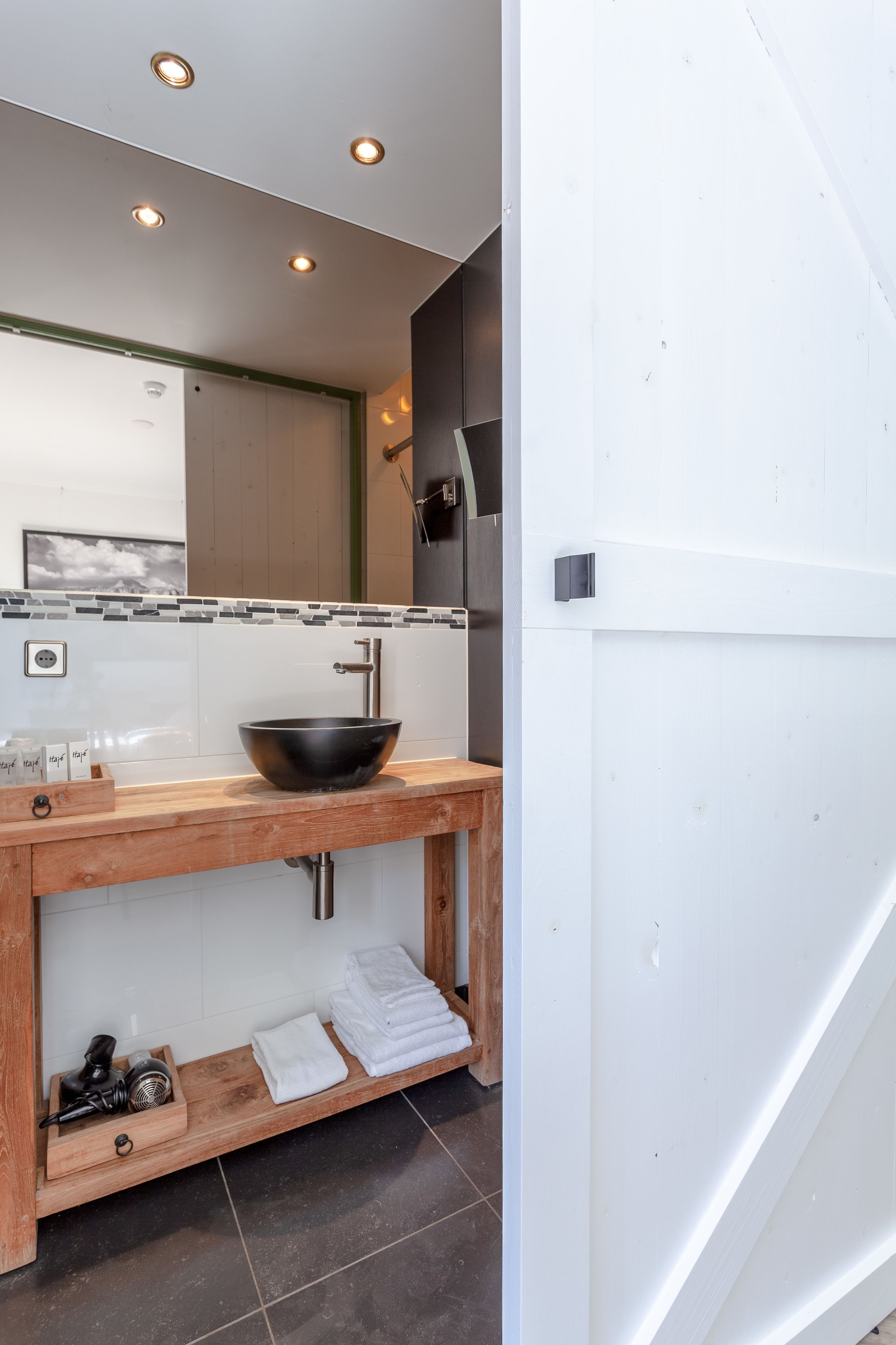 Uma casa de banho moderna com um lavatório de madeira, uma lavatório escultural preta e um espelho em cima. Há toalhas e artigos de higiene pessoal no lavatório.