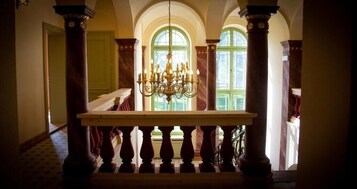 Um amplo salão com lustre, janelas em arco e colunas clássicas.