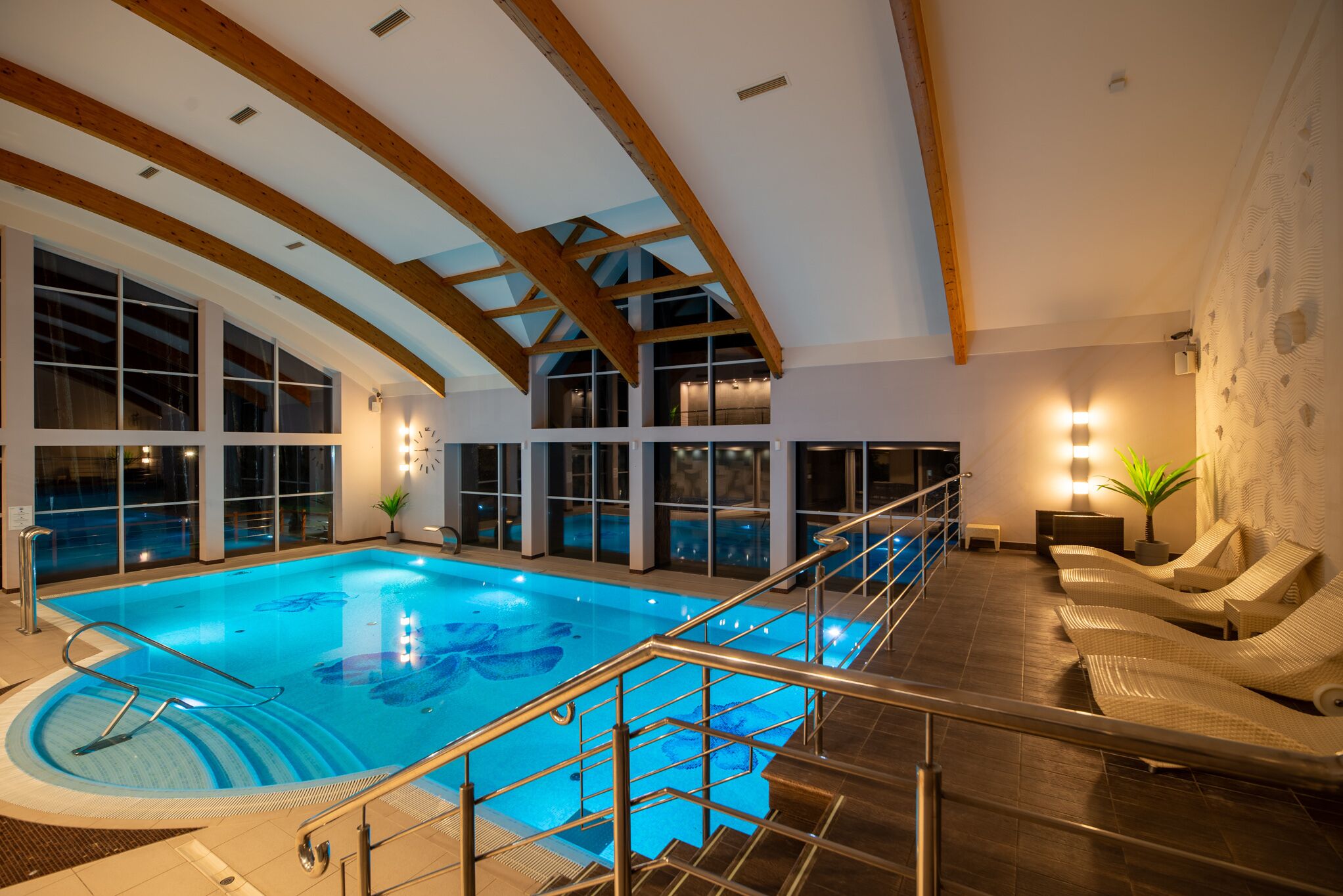 Piscina interna com teto alto e vigas de madeira, cercada por cadeiras de descanso e grandes janelas.