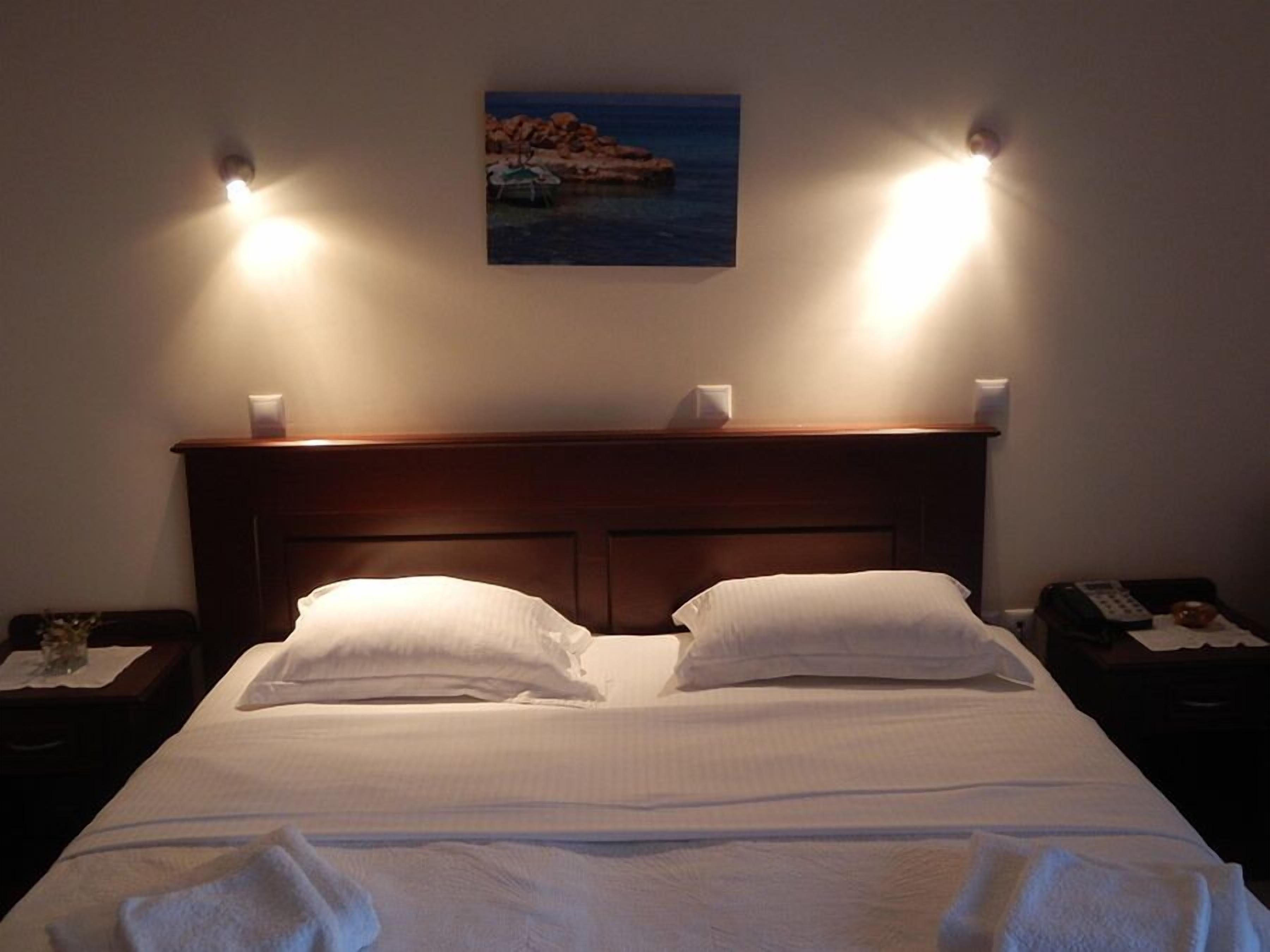 Quarto de hotel com uma cama, duas mesas de cabeceira, um criado-mudo, um quadro na parede e duas luzes de parede.