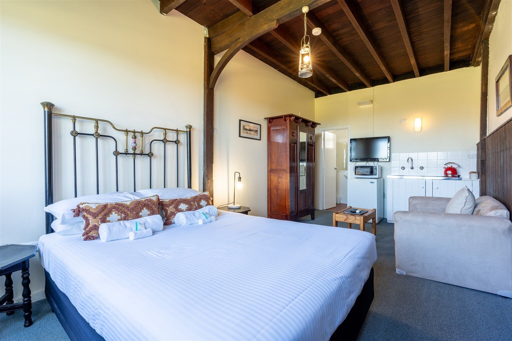Quarto com cama grande, guarda-roupa de madeira, mesa pequena e uma kitchenette ao fundo.