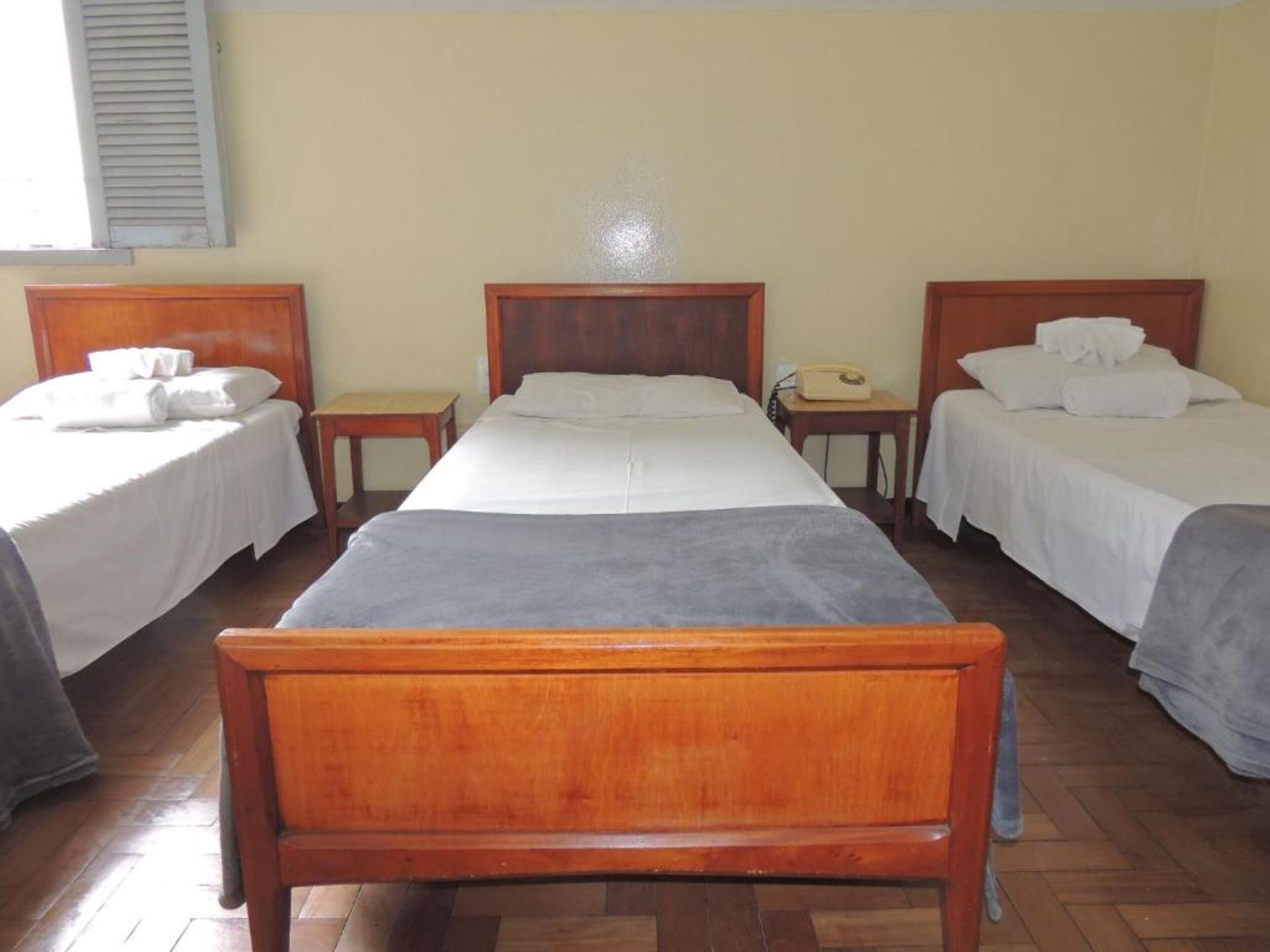 Quarto de hotel com três camas de solteiro, cabeceiras de madeira, e uma cama central com manta cinza.