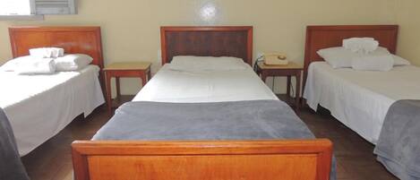 Quarto de hotel com três camas de solteiro, cabeceiras de madeira, e uma cama central com manta cinza.