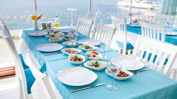 Mesa posta para refeição, com vista para o mar e barcos ao fundo.