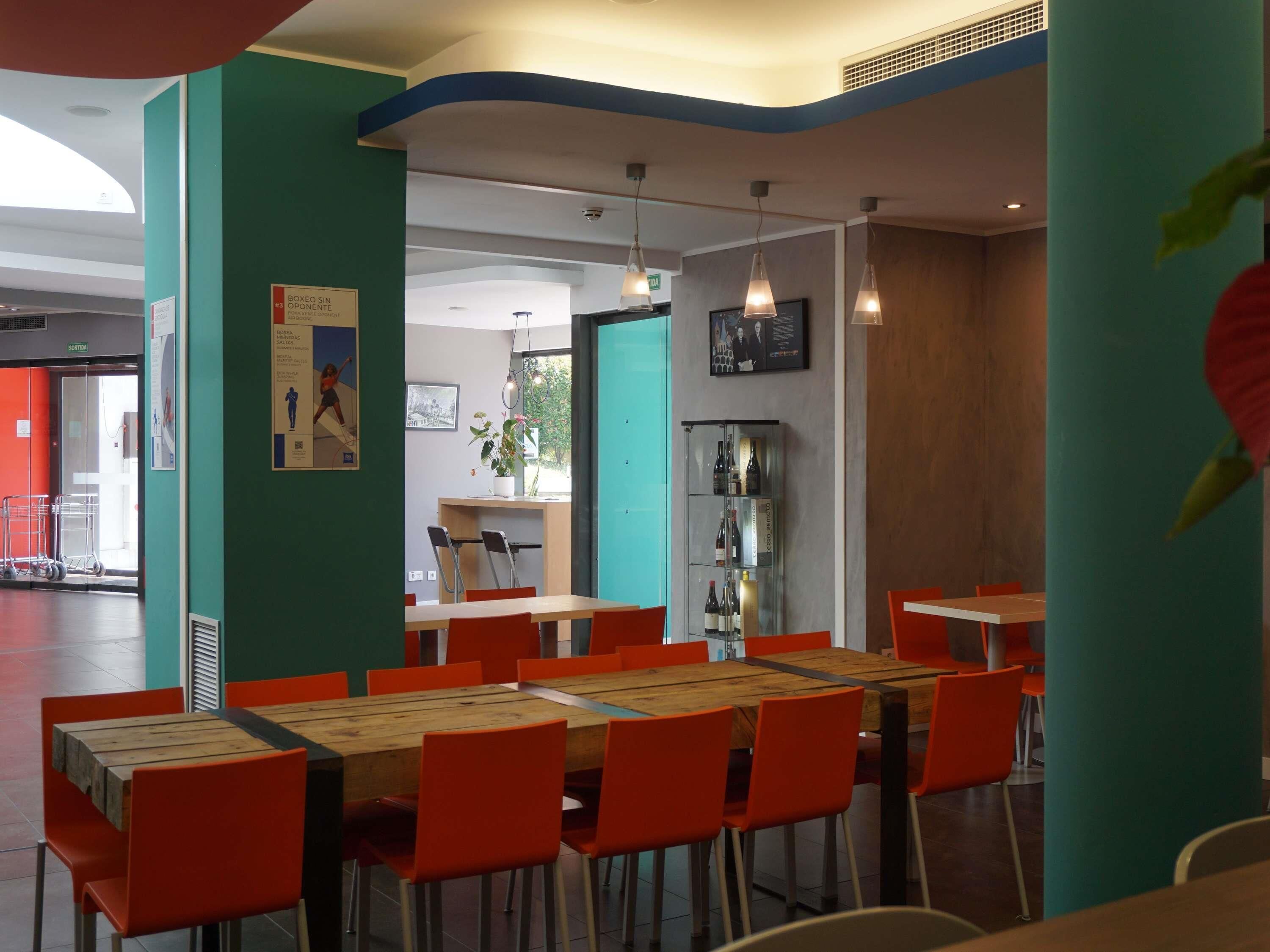 Restaurante com uma longa mesa de madeira e cadeiras laranjas, uma vitrine de vidro com garrafas e uma parede azul de destaque.