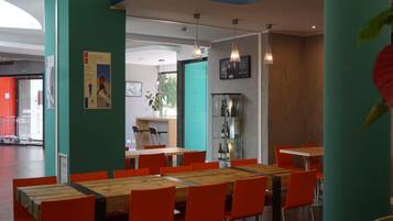 Restaurante com uma longa mesa de madeira e cadeiras laranjas, uma vitrine de vidro com garrafas e uma parede azul de destaque.