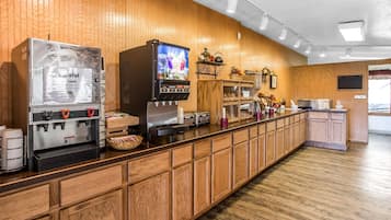 Uma estação de café por autoatendimento, com um dispensador de bebidas, uma tela de televisão e diversos alimentos no balcão.