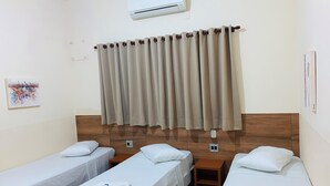 Quarto de hotel com três camas de solteiro, cabeceira de madeira, ar condicionado, tomada e criado-mudo pequeno.