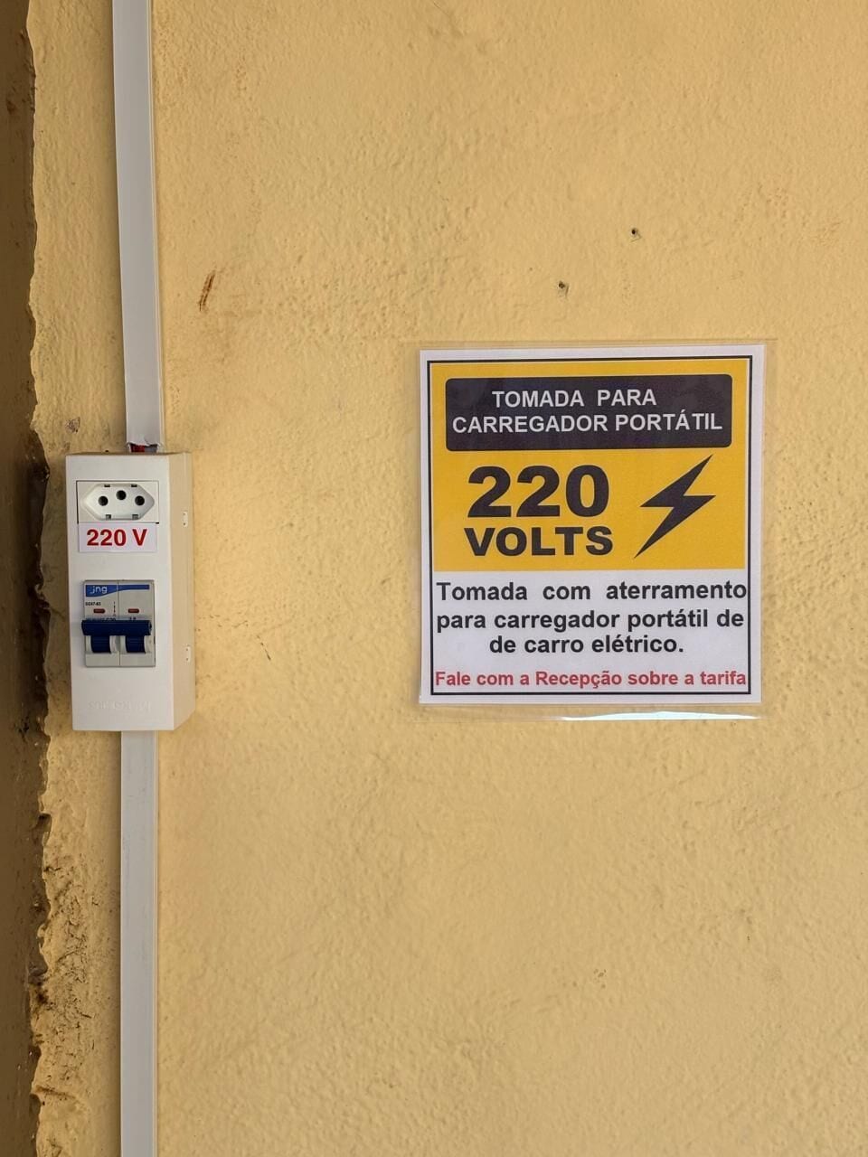 Caixa elétrica fixada na parede com uma placa indicando tomada de 220 volts para carregadores portáteis de veículos elétricos.