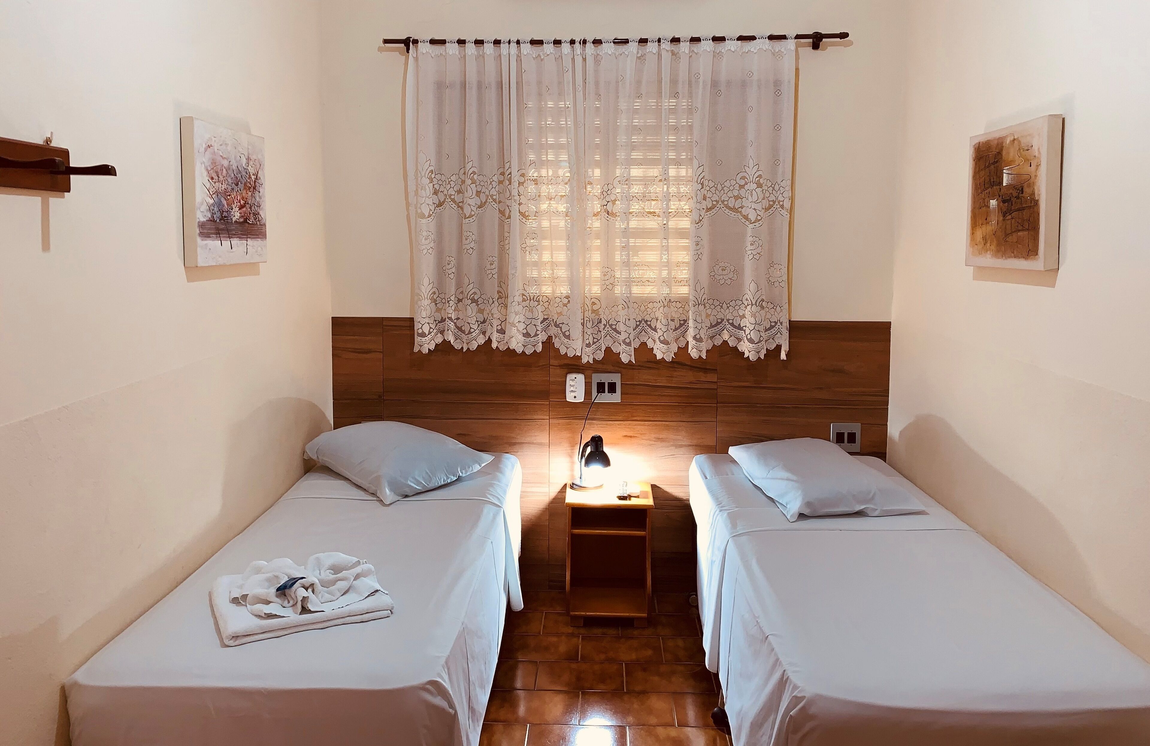 Quarto com duas camas, mesa de cabeceira com abajur e obras de arte nas paredes.