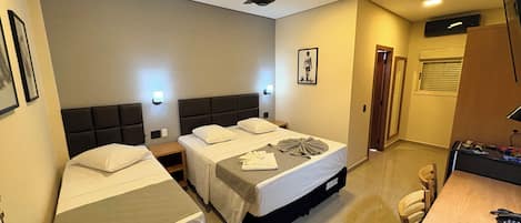 Quarto de hotel com duas camas, uma escrivaninha, uma cadeira, um ventilador de teto e uma porta que dá para outro cômodo.