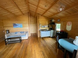 Interior de uma cabana de madeira com sofá, mesa de jantar, cozinha compacta e um quadro na parede.