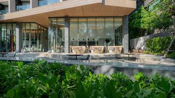 Um edifício moderno com fachada de vidro, área de estar externa com móveis de vime e uma piscina.