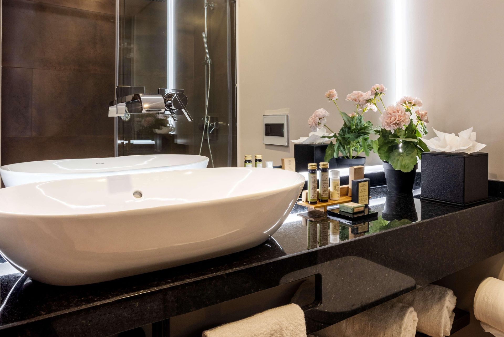 Um banheiro moderno com uma pia oval grande, box de vidro e bancada com produtos de higiene pessoal e um vaso de flores.
