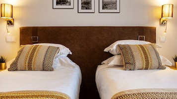 Quarto de hotel com duas camas, cada uma com almofadas estampadas e cabeceira marrom. Há luminárias fixadas na parede, uma de cada lado da cama.
