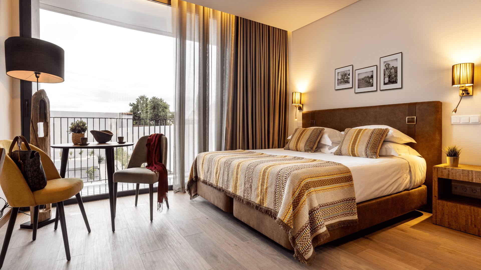 Quarto de hotel com uma cama grande, varanda com mesa e cadeiras, e vista para o exterior.