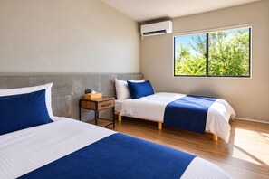 Quarto de hotel com duas camas, uma mesinha de cabeceira, ar condicionado e janela vista para vegetação.