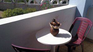Uma pequena mesa ao ar livre com um vaso de flores secas, duas cadeiras de plástico e uma parede branca com um muro decorativo ao fundo.