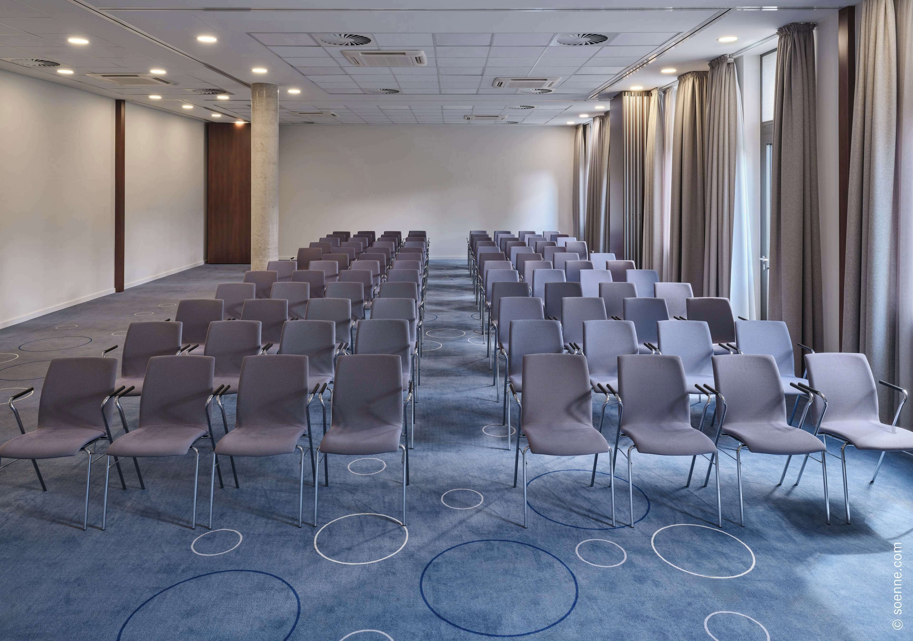 Uma sala de conferências com fileiras de cadeiras cinzas, um carpete com padrões circulares e janelas amplas com cortinas.