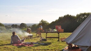 Um camping com uma tenda, uma mesa com utensílios de cozinha, e pessoas e um cão aproveitando a natureza.