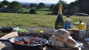 Mesa posta para piquenique com salsichas, salada, pão, acompanhamentos e uma garrafa de vinho, com vista panorâmica ao fundo.