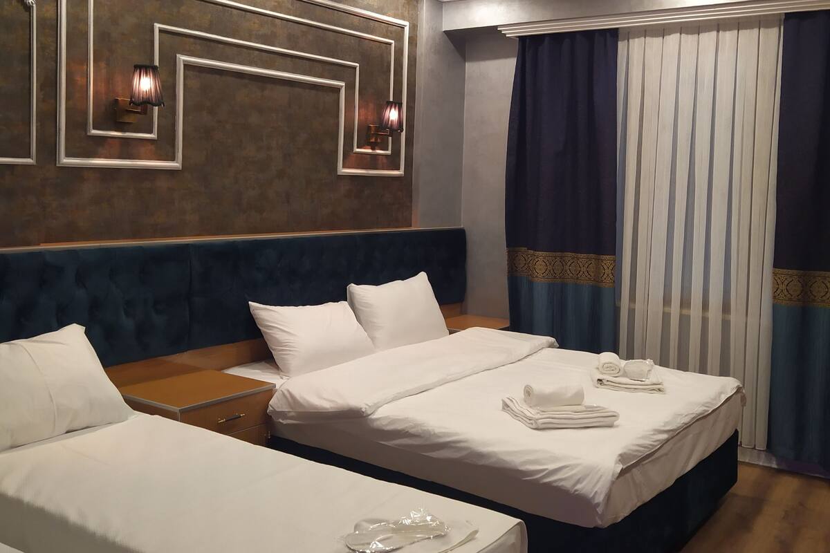 Quarto de hotel com duas camas, cabeceira de madeira, luminária fixada na parede e janela com cortinas.