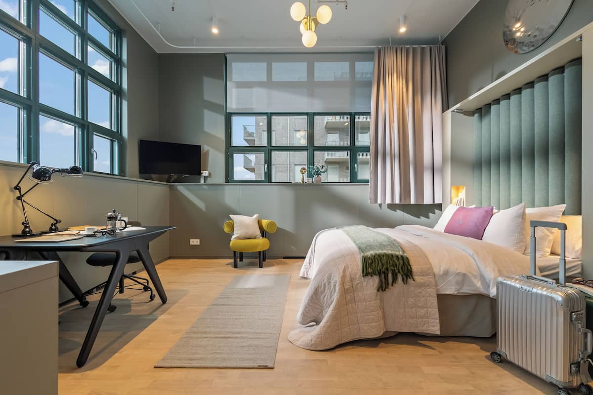 Quarto de hotel moderno com uma cama grande, uma mesa com computador, uma cadeira e uma janela com cortinas.