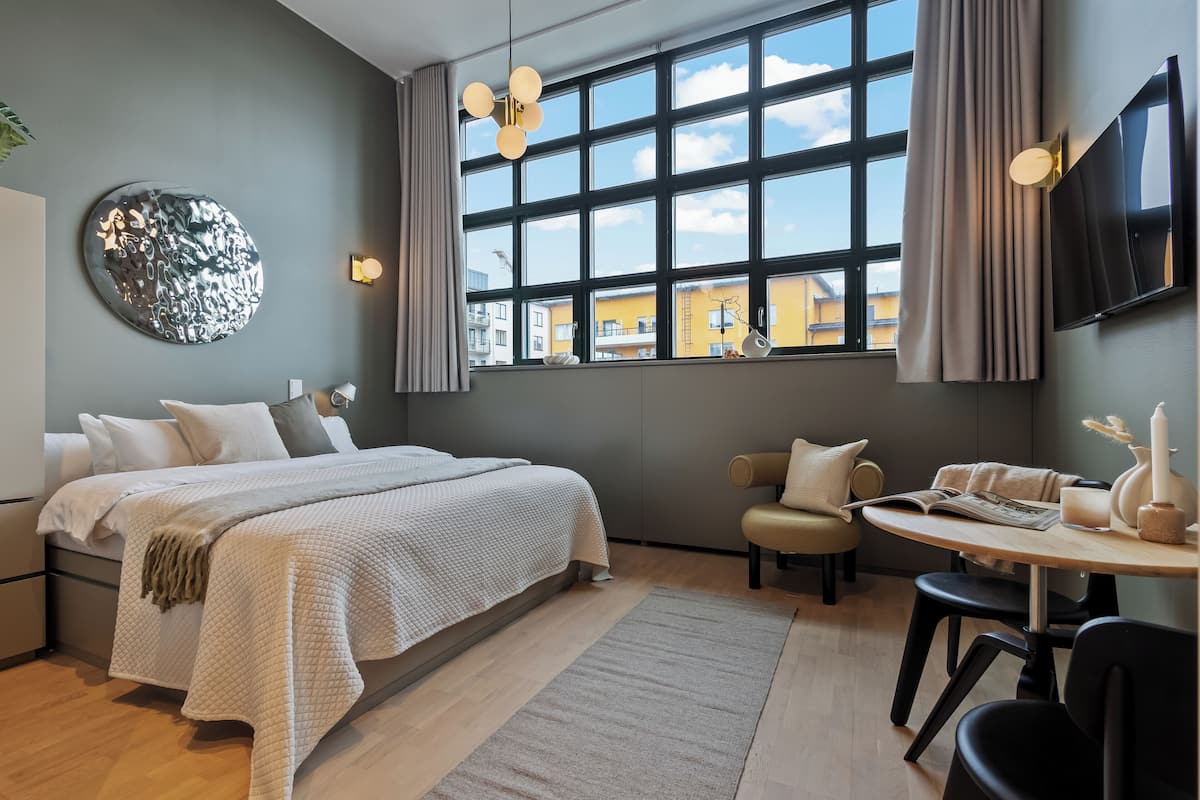 Quarto de hotel moderno com uma janela grande, uma cama, uma mesa de trabalho e uma televisão.