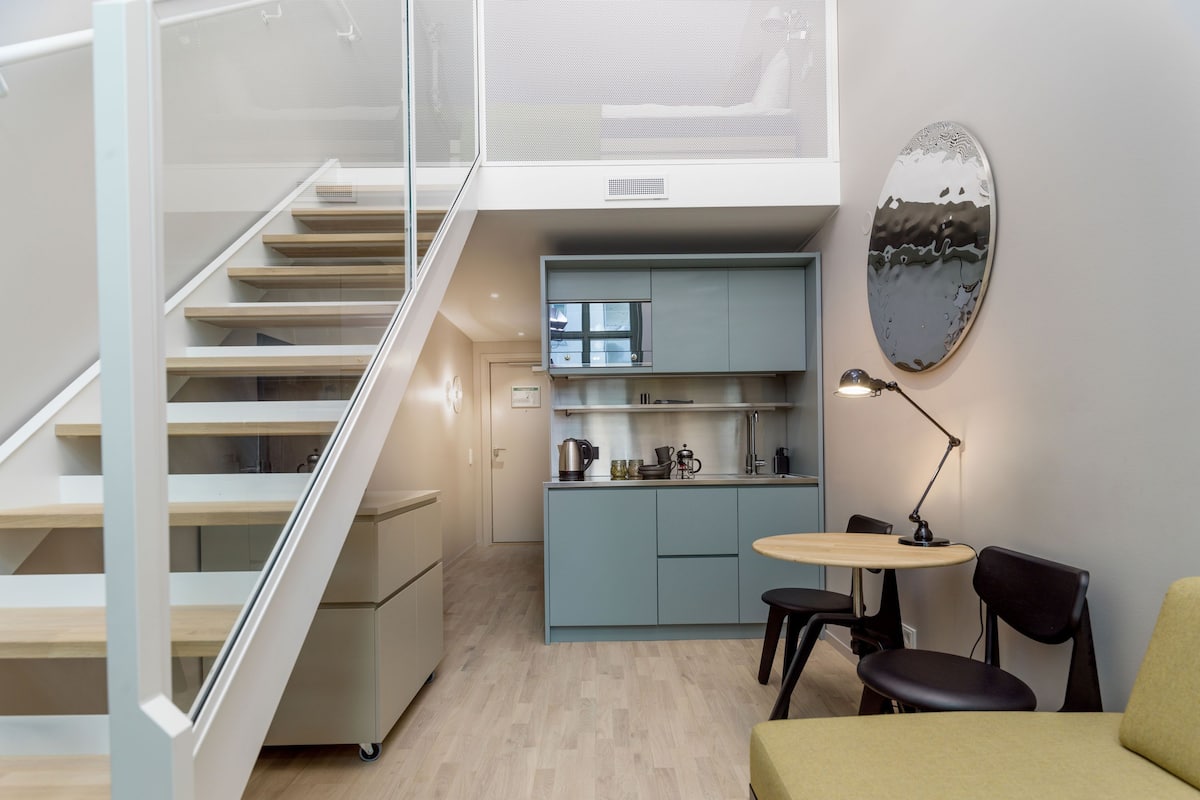 Um interior moderno com escada que leva a um mezanino, cozinha com armários azul-claros, mesa redonda com cadeiras e luminária fixada na parede.