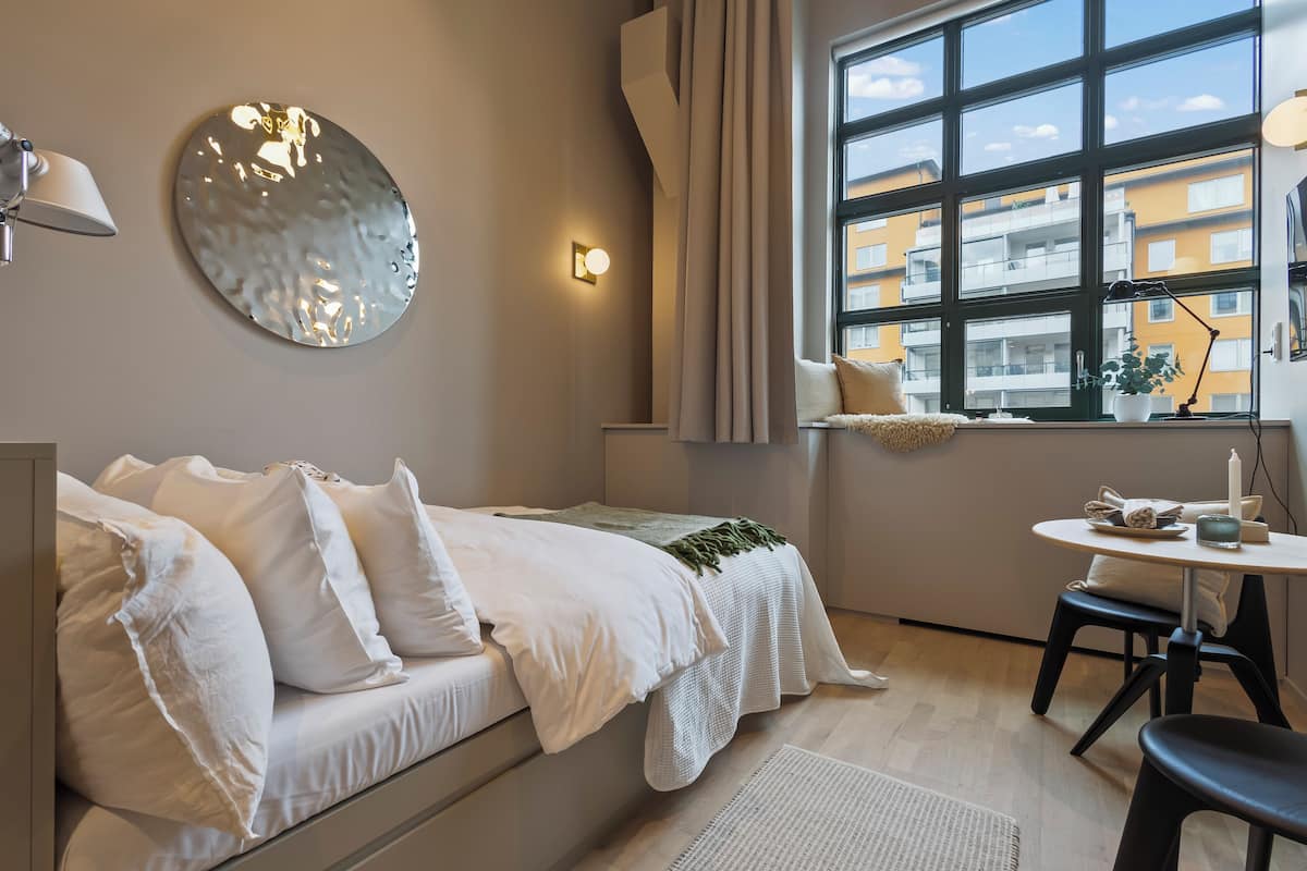 Quarto moderno com cama, decoração circular na parede, janela com cortinas e uma pequena mesa com abajur.