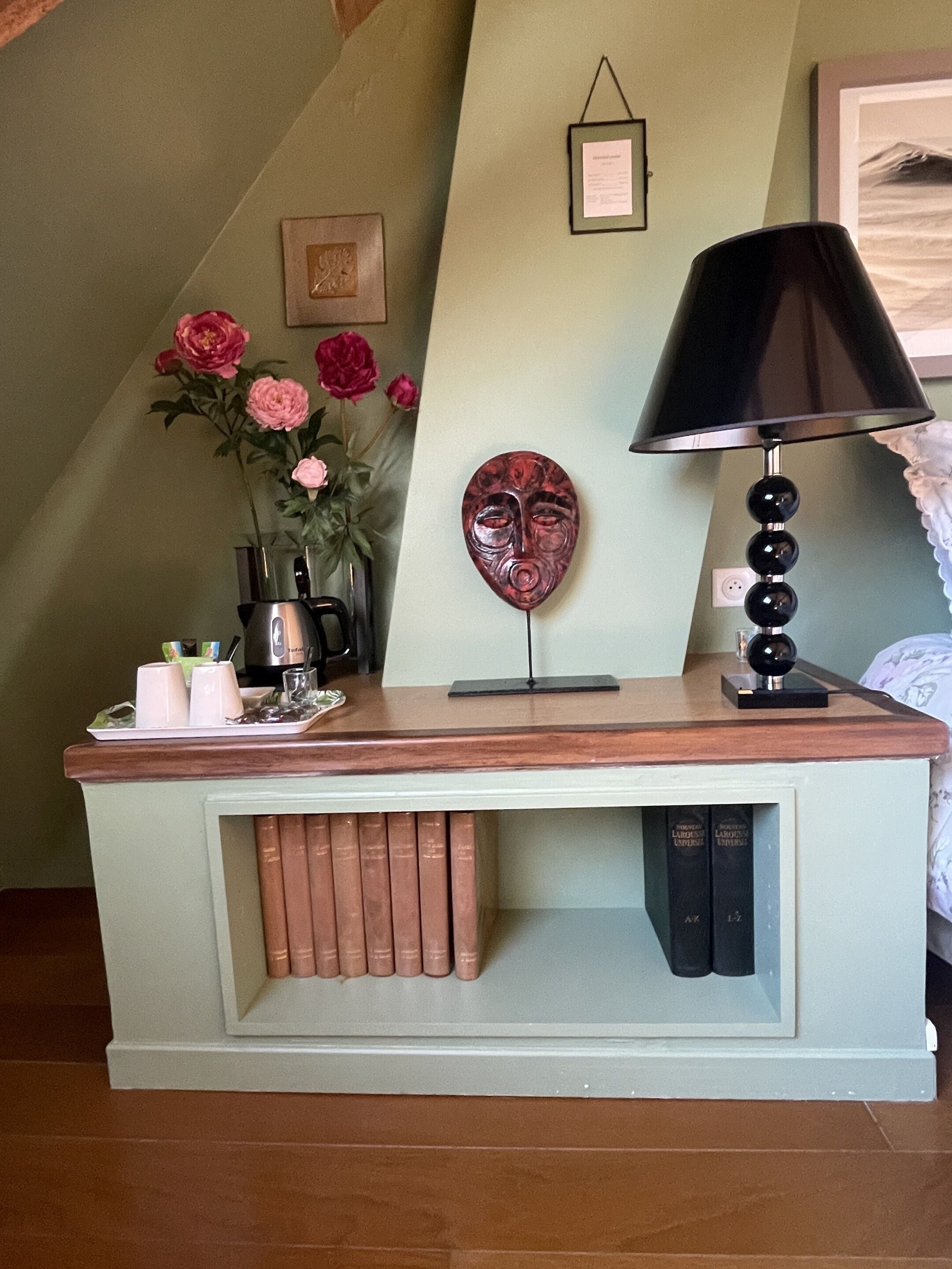 Um quarto pequeno e acolhedor com uma escrivaninha de madeira, um vaso de flores, uma máscara decorativa e um abajur.
