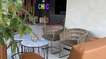 Uma área de estar externa com sofás de couro e mesas redondas, um letreiro de neon com a inscrição "MODERN AND CHIC" e uma planta em primeiro plano.