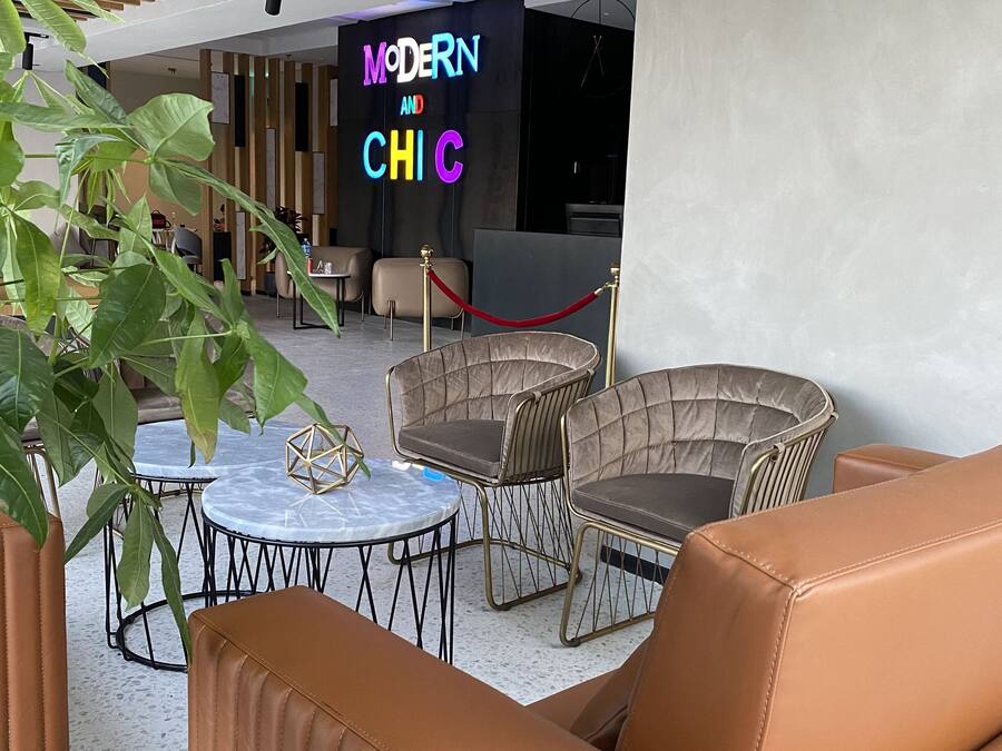 Uma área de estar externa com sofás de couro e mesas redondas, um letreiro de neon com a inscrição "MODERN AND CHIC" e uma planta em primeiro plano.