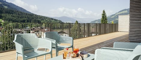 Terraço com duas cadeiras de metal e uma mesa, um tabuleiro com bebidas e petiscos, e vista para montanhas.