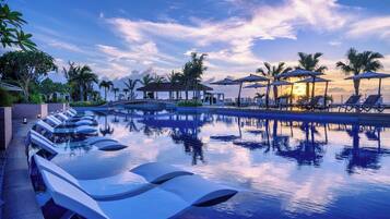 Piscina com espreguiçadeiras e guarda-sóis, cercada por palmeiras e um pôr do sol.